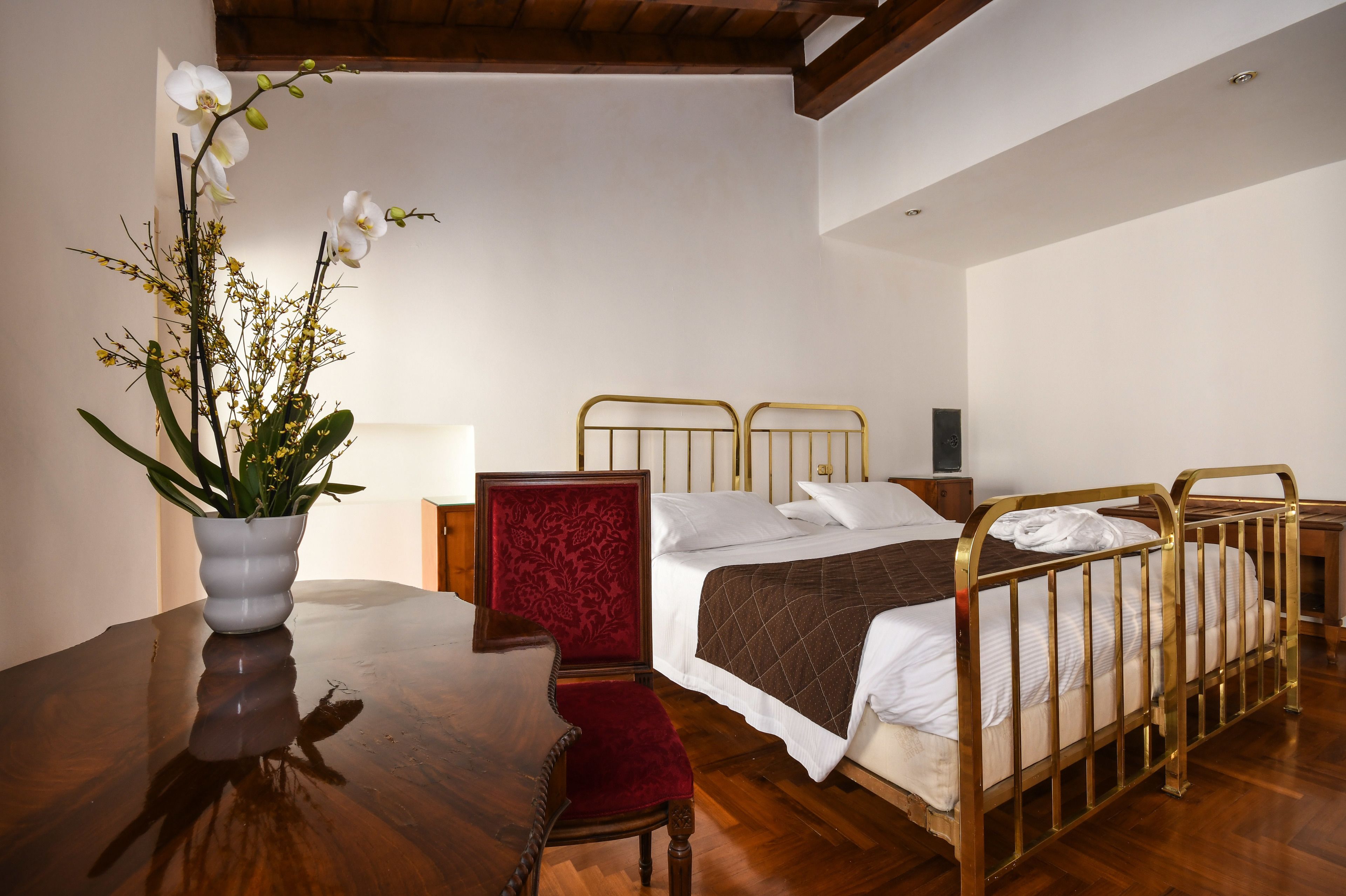 Quarto de hotel com piso de madeira, cama com estrutura metálica, poltrona vermelha estofada, mesa de madeira e um vaso com flores brancas.