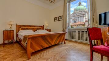 Quarto de hotel com uma cama de madeira, uma cadeira vermelha, uma televisão e vista para um edifício com uma cúpula através de uma janela.