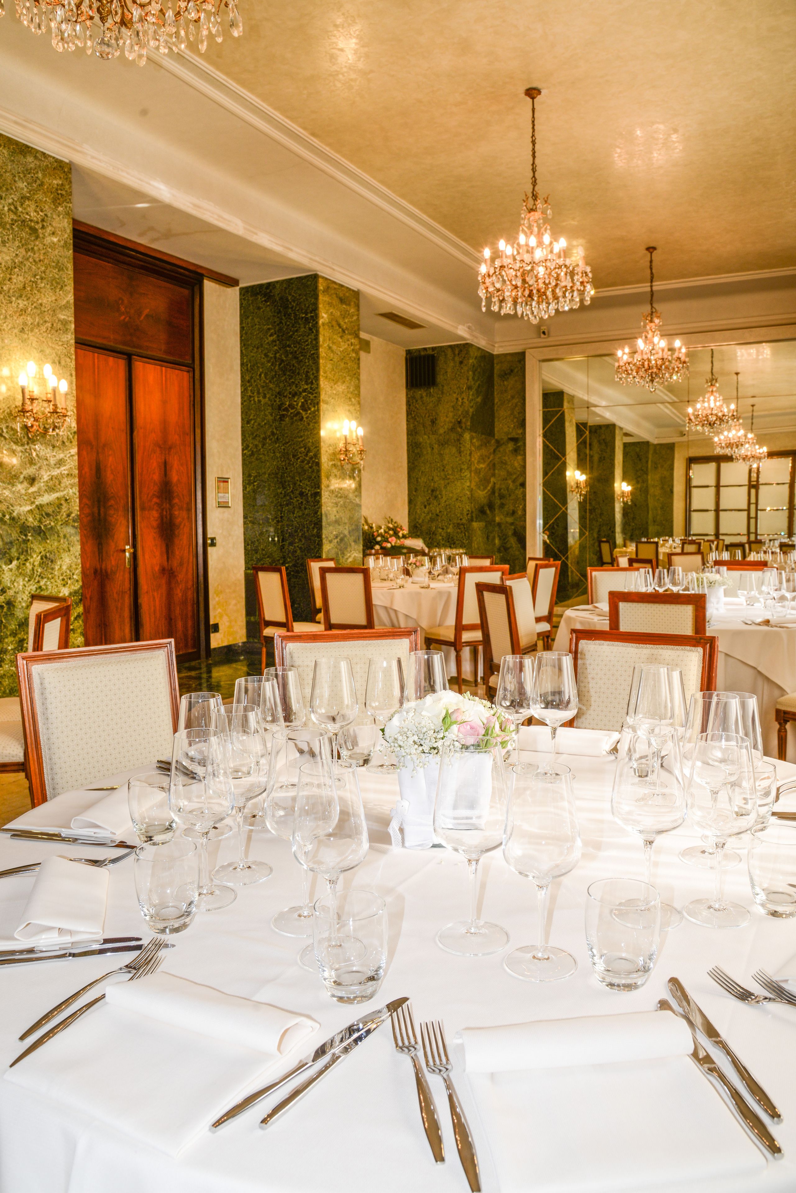 Uma sala de banquetes com várias mesas preparadas para refeições, com lustres e paredes de mármore.