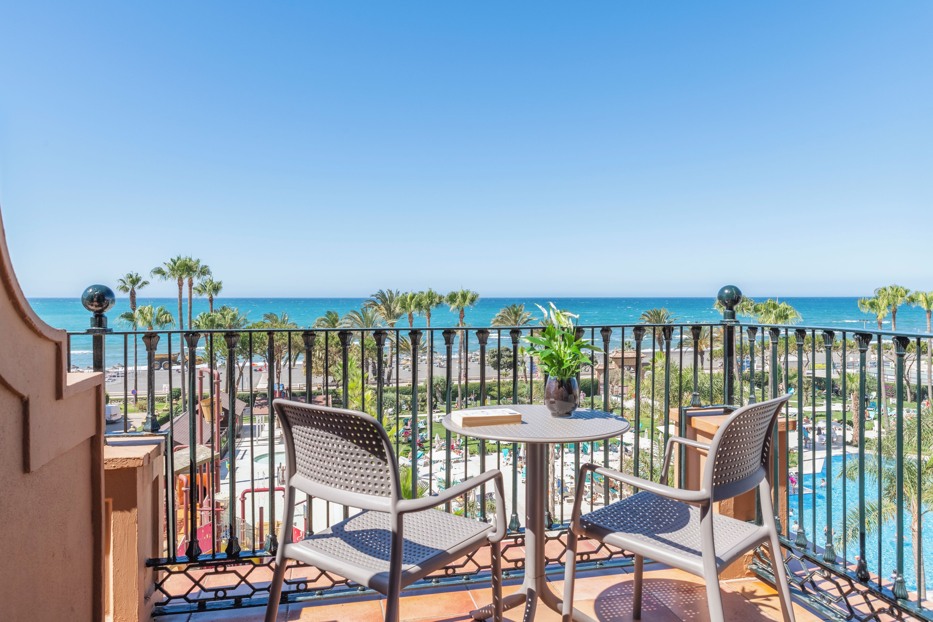 Varanda com duas cadeiras e uma mesa pequena, com vista para uma piscina e uma praia.