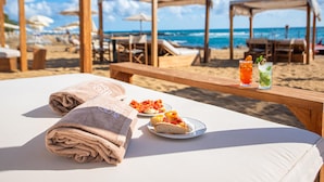 Um espaço de relaxamento à beira-mar, com espreguiçadeiras, uma mesa de madeira com bebidas e vista para o oceano.