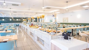 Uma cafeteria ampla com várias mesas e cadeiras, um balcão de buffet com diversos alimentos e luminárias pendentes.