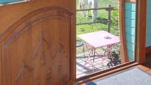 Uma porta de madeira com entalhes elaborados, vista de um cavalo branco pastando em área externa com uma mesa e cadeiras cor de rosa, e um candeeiro pendurado na parede.