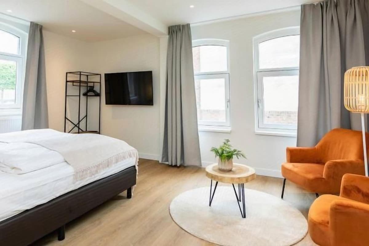 Quarto de hotel com cama, televisão, uma mesinha com uma planta e duas cadeiras.