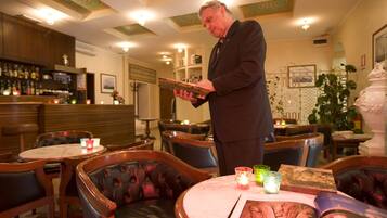 Um homem lendo um livro em um quarto bem iluminado, com poltronas de couro, um bar ao fundo e teto decorativo.