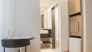Quarto de hotel moderno com piso de madeira, uma pequena mesa redonda com uma cadeira, uma luminária de teto branca e vista para o banheiro.