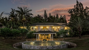 Uma casa com alpendre, um lago circular com nenúfares e um jardim bem cuidado, com diversas árvores e arbustos.