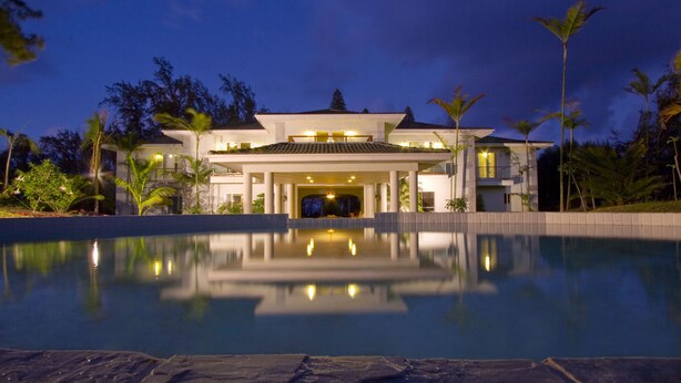 Uma casa grande e moderna com piscina em frente, rodeada por vegetação tropical.