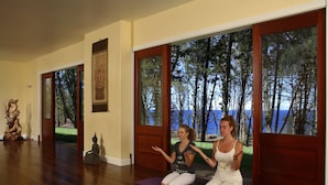 Duas pessoas praticando yoga em um espaço com piso de madeira e grandes janelas com vista para árvores e um corpo d'água.