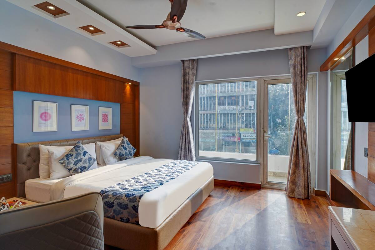 Quarto de hotel moderno com uma cama grande, cabeceira de madeira, ventilador de teto e uma janela ampla com cortinas.