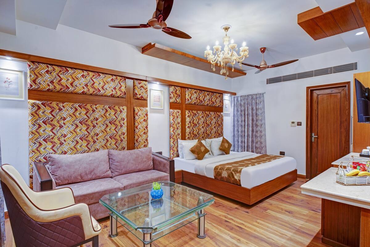 Quarto de hotel com uma cama, um sofá, uma mesa de centro de vidro, um ventilador de teto, um lustre e painéis de madeira nas paredes.