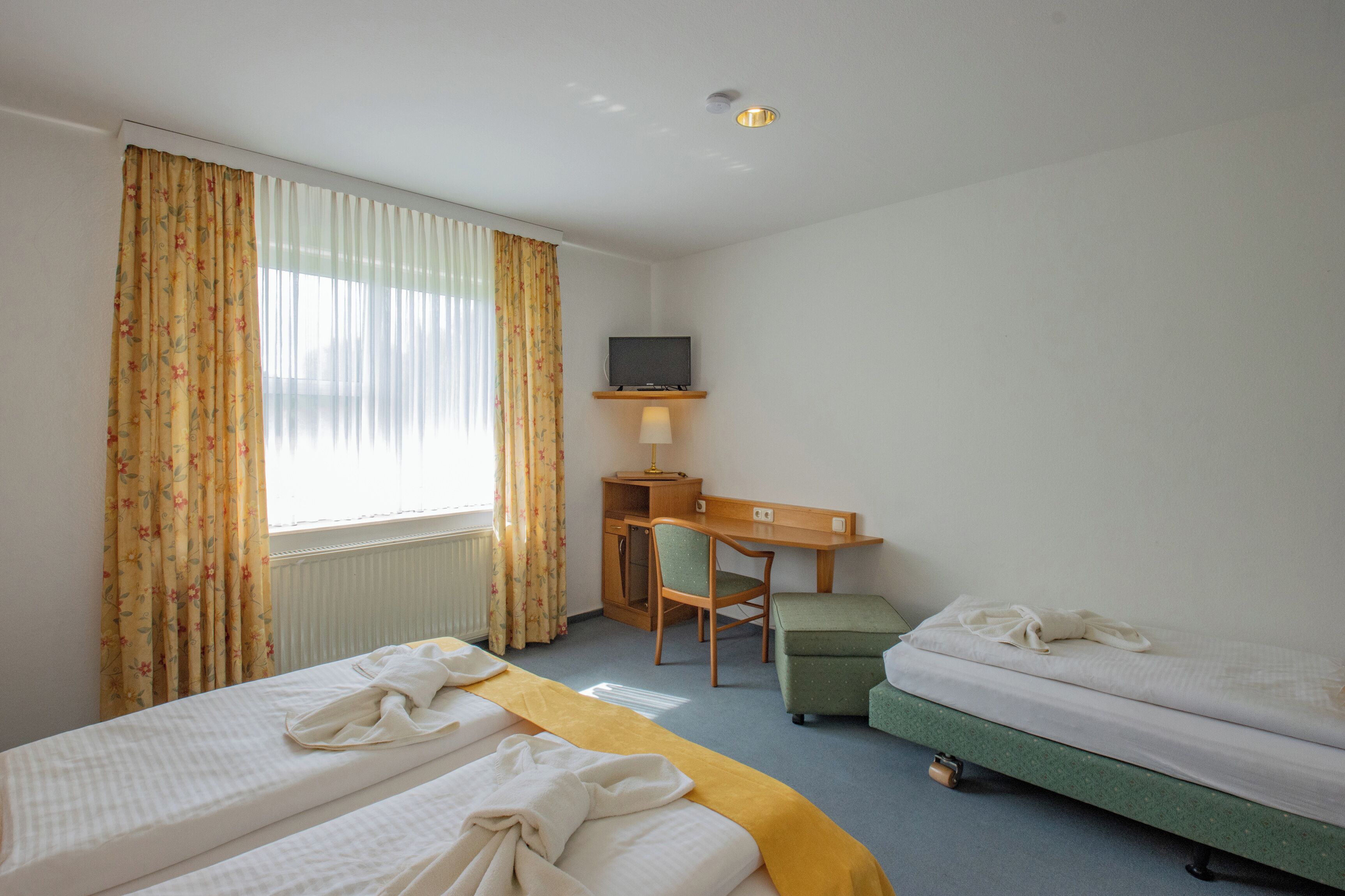 Quarto de hotel com duas camas, uma mesa, uma cadeira, televisão e janela com cortinas.