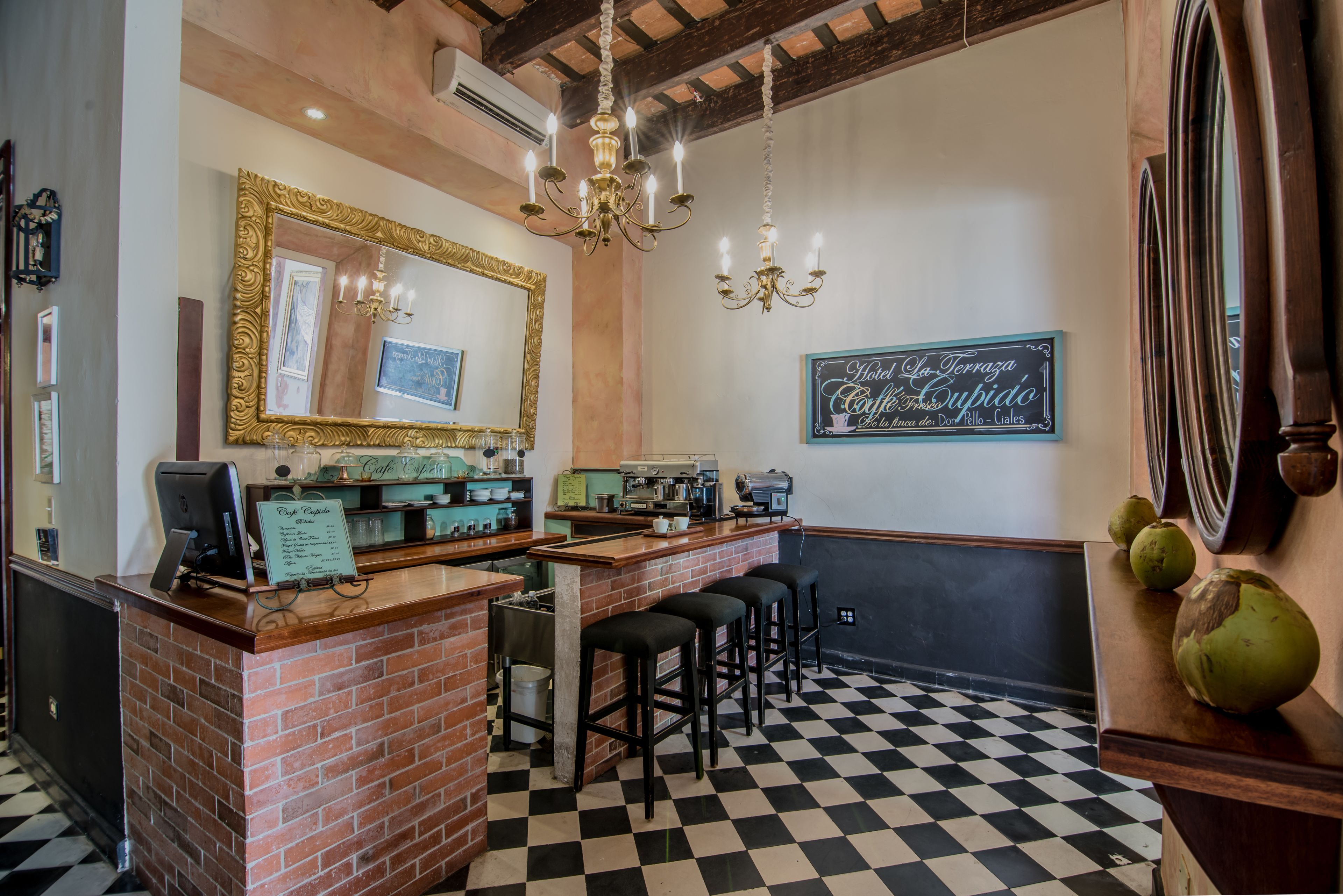 Uma área de bar com piso xadrez, balcão com banquetas, espelho e um letreiro indicando cardápio de café e sobremesas.