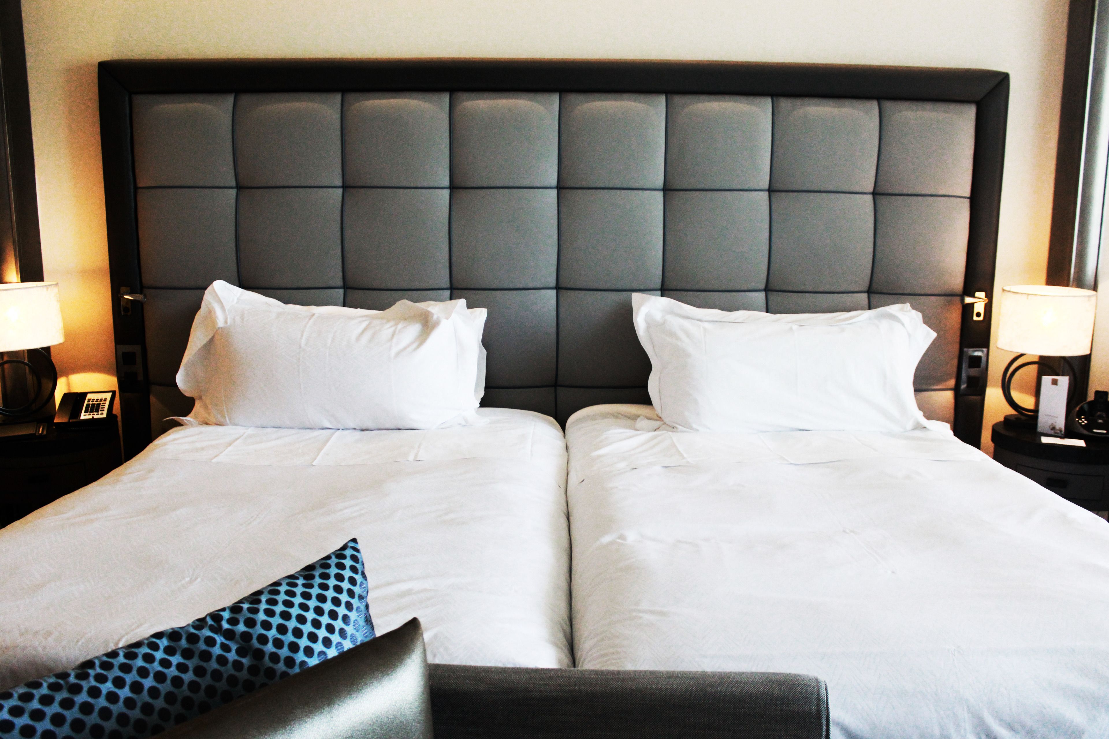 Quarto de hotel com duas camas, cada uma com roupa de cama branca e cabeceira escura. Há duas mesas de cabeceira com luminárias e um pequeno relógio em cada uma.
