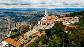 Um edifício grande e histórico, com uma torre imponente, rodeado por vegetação exuberante e uma paisagem urbana ao fundo.