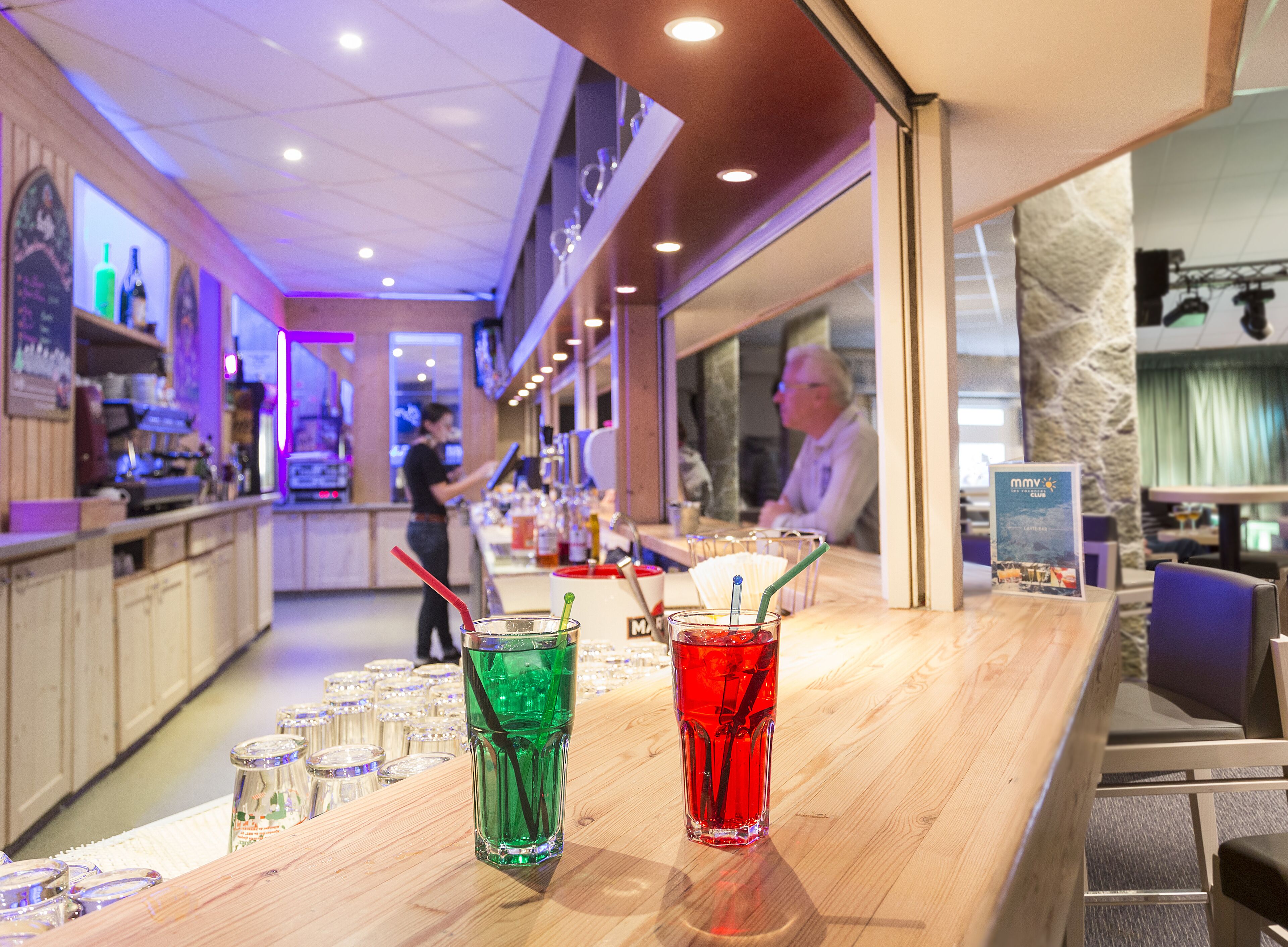 Uma área de bar com duas bebidas no balcão, um bartender e um cliente ao fundo.