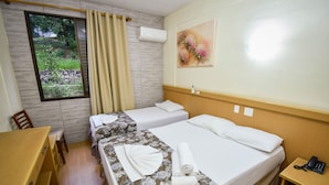 Quarto de hotel com duas camas, uma escrivaninha de madeira, uma cadeira, janela com vista para vegetação e um quadro na parede.