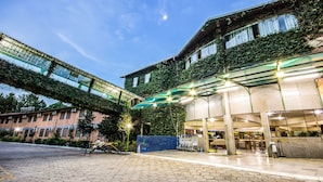 Entrada de hotel com área externa coberta, fachada de vidro e uma ponte sobre o estacionamento.