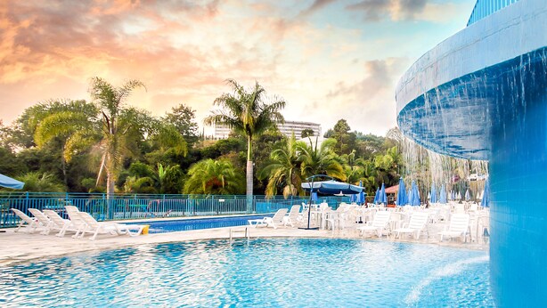 Área de piscina com espreguiçadeiras e guarda-sóis, cercada por palmeiras e um edifício moderno.