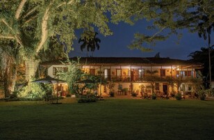 Um edifício de dois andares com varanda, rodeado por árvores e um gramado bem cuidado.