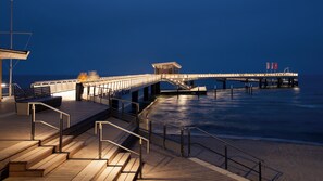Um deck de madeira com degraus que levam a um cais que se estende sobre a água, um bote salva-vidas atracado no final, e uma estação de bote salva-vidas com um pequeno edifício.