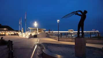 Um calçadão à beira-mar com uma estátua, um píer e um edifício com mesas ao ar livre.