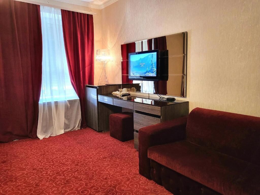 Quarto de hotel com tapete vermelho, escrivaninha com computador, televisão e sofá vermelho.