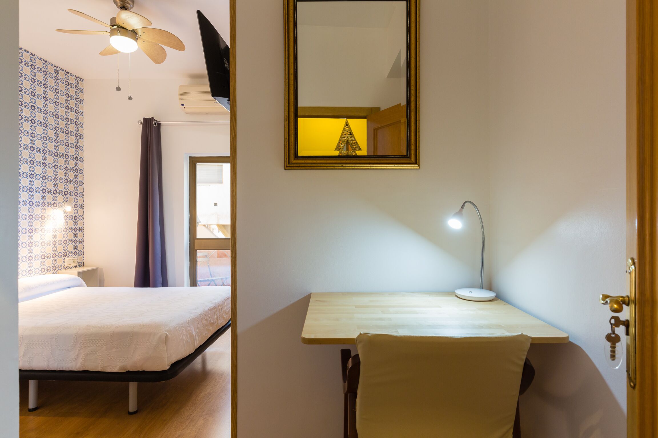 Quarto com cama, escrivaninha com cadeira, ventoinha no teto, televisão e espelho.