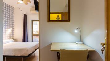 Quarto com cama, escrivaninha com cadeira, ventoinha no teto, televisão e espelho.