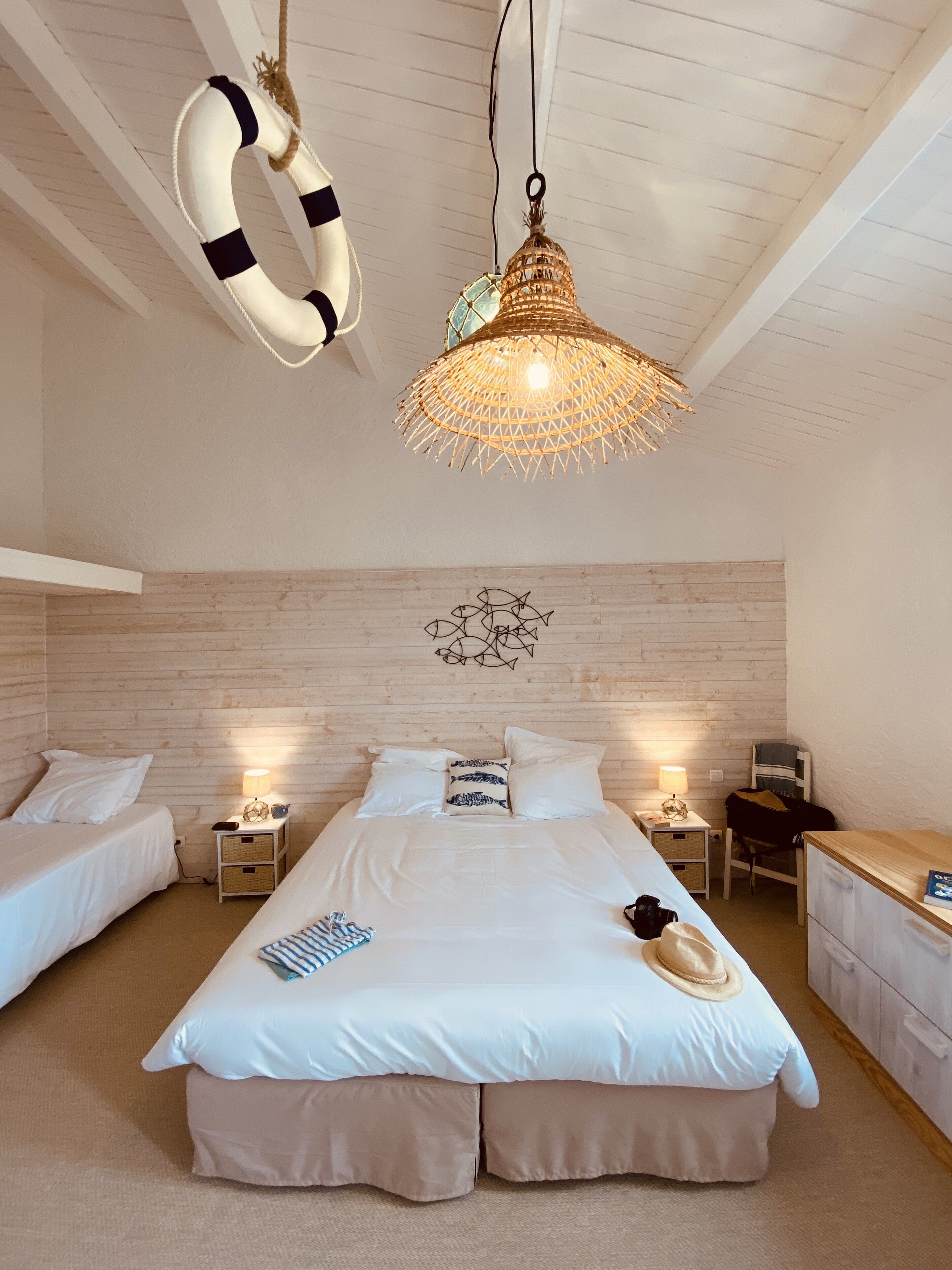 Quarto com uma cama grande, duas mesas de cabeceira, um salva-vidas pendurado e uma luminária de tecido.