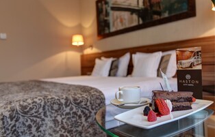 Quarto de hotel com uma cama, uma mesa de vidro com um prato de sobremesa, uma chávena de café e uma moldura na parede.