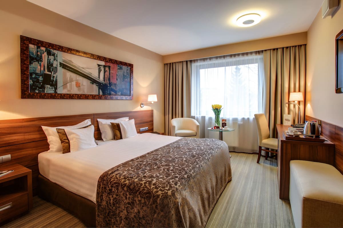 Quarto de hotel com uma cama grande, uma escrivaninha, uma cadeira, uma mesa pequena com abajur e um quadro na parede.