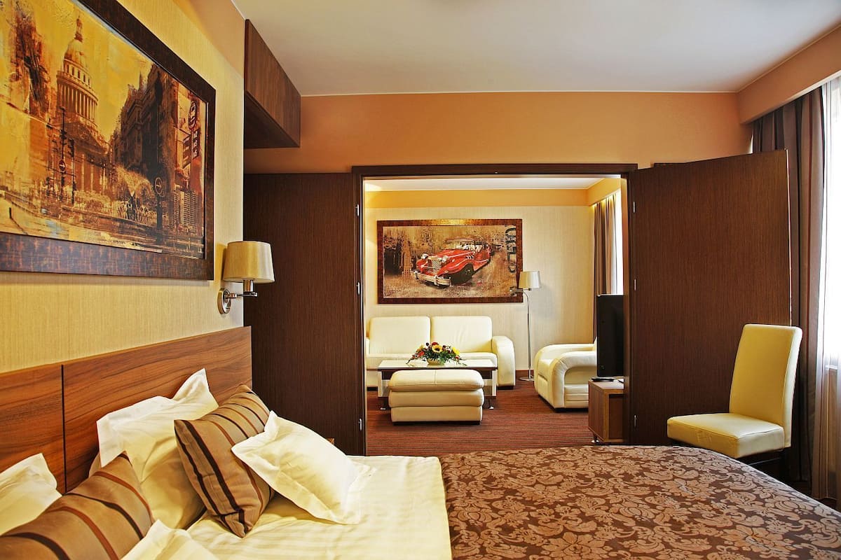 Quarto de hotel com uma cama grande, um sofá, uma cadeira e uma televisão. Há quadros nas paredes e um abajur na mesa lateral.