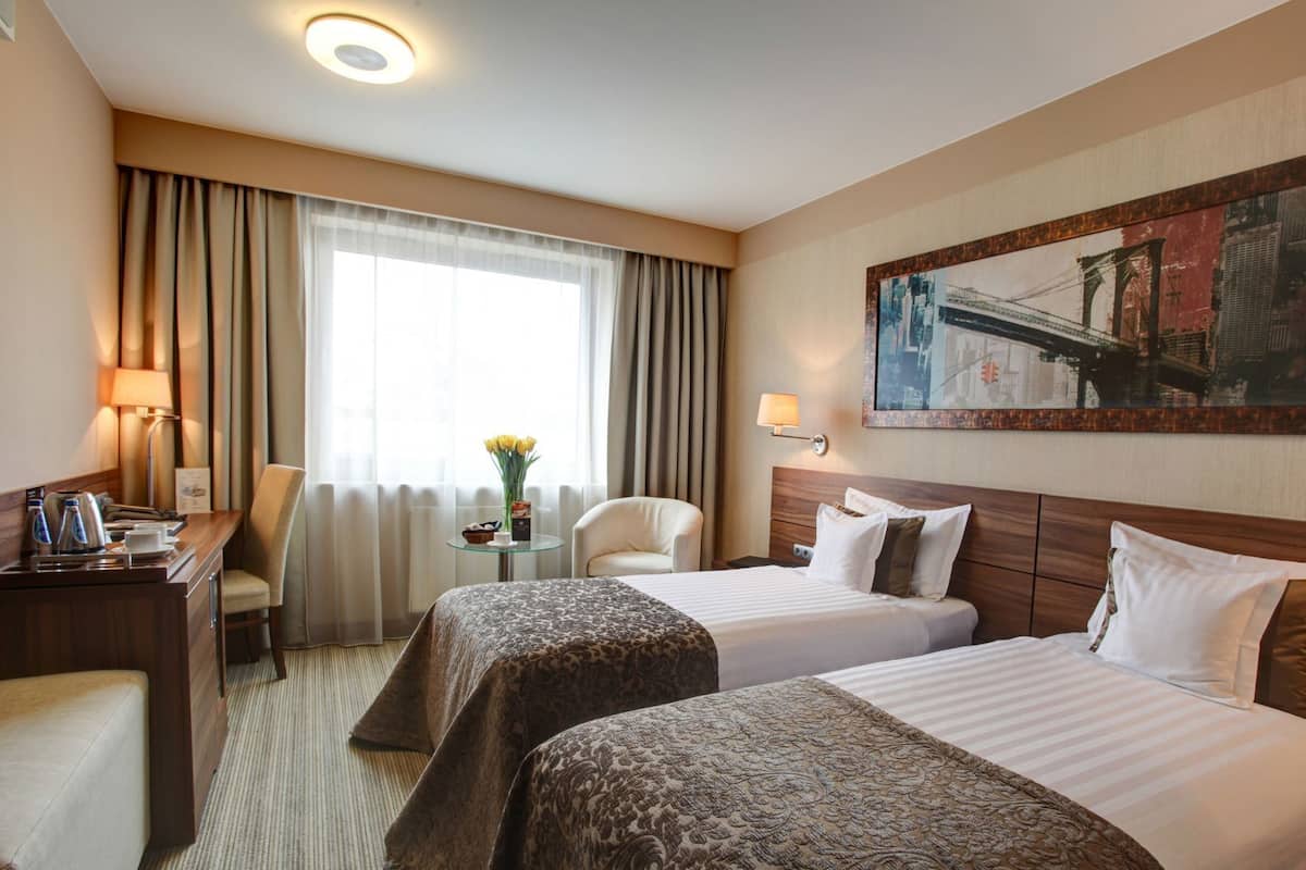 Quarto de hotel com duas camas, uma escrivaninha, uma cadeira, um abajur e um quadro na parede.