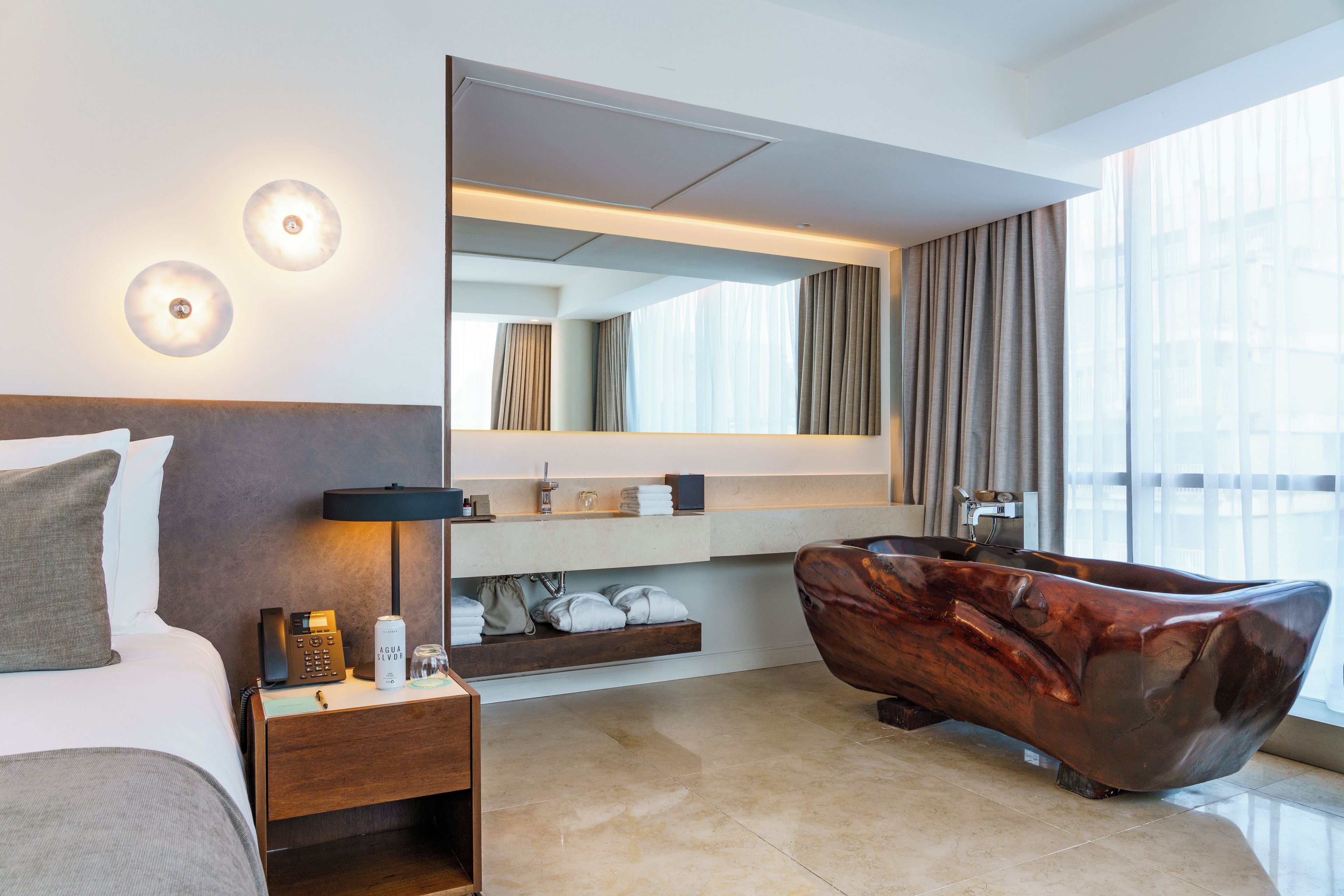 Quarto de hotel moderno com uma cama grande, uma toucador com espelho, uma banheira e uma janela ampla com cortinas.