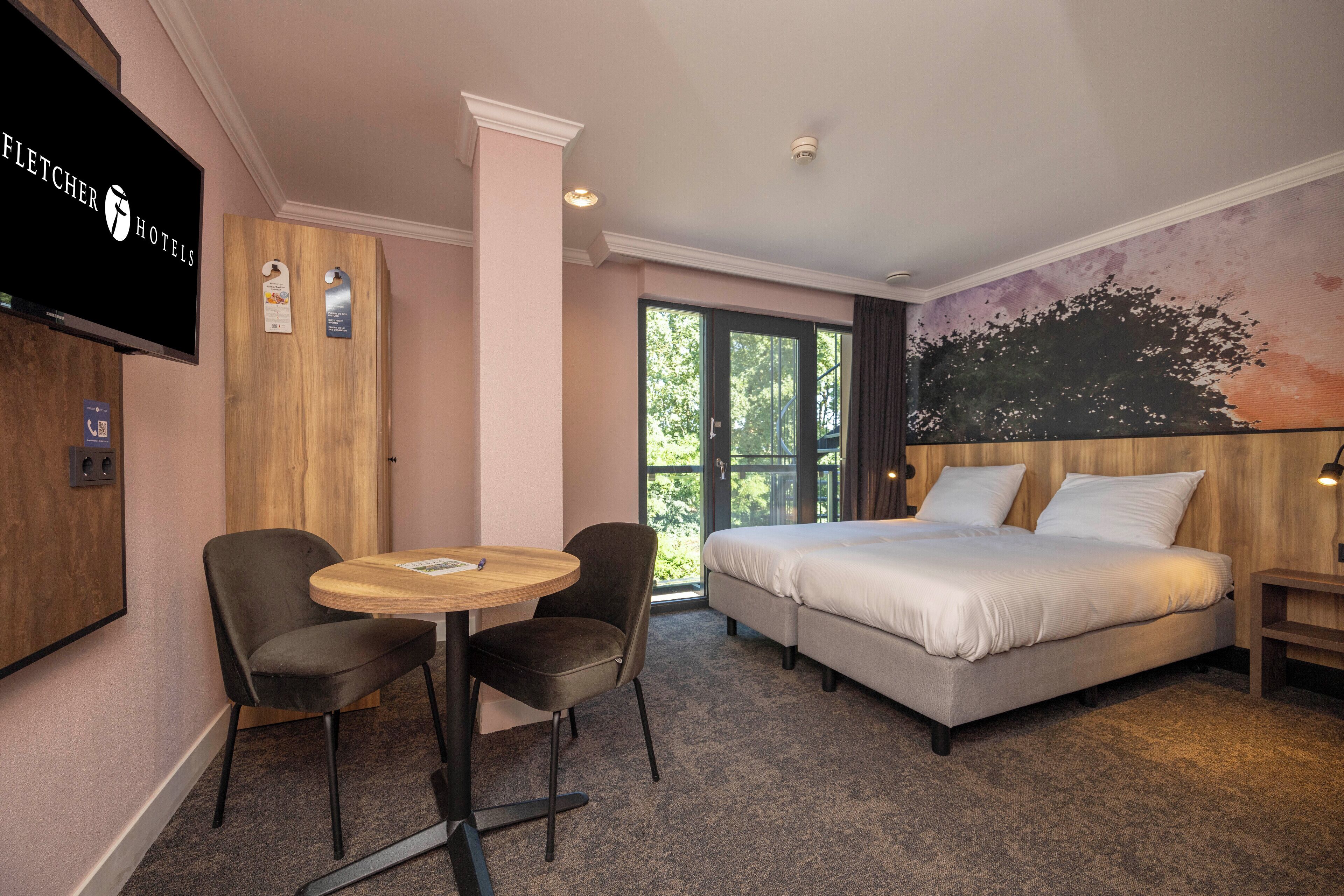 Quarto de hotel com cama, duas cadeiras, uma mesa pequena, televisão e uma janela grande.