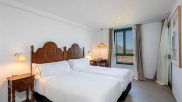Uma cama de casal com cabeceira de madeira, duas mesas de cabeceira com luminárias, vista do exterior pela janela e uma pequena mesa com uma cadeira.