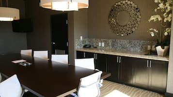 Uma sala de conferências moderna com uma mesa de madeira escura, cadeiras brancas, bancada de granito e um espelho decorativo na parede.