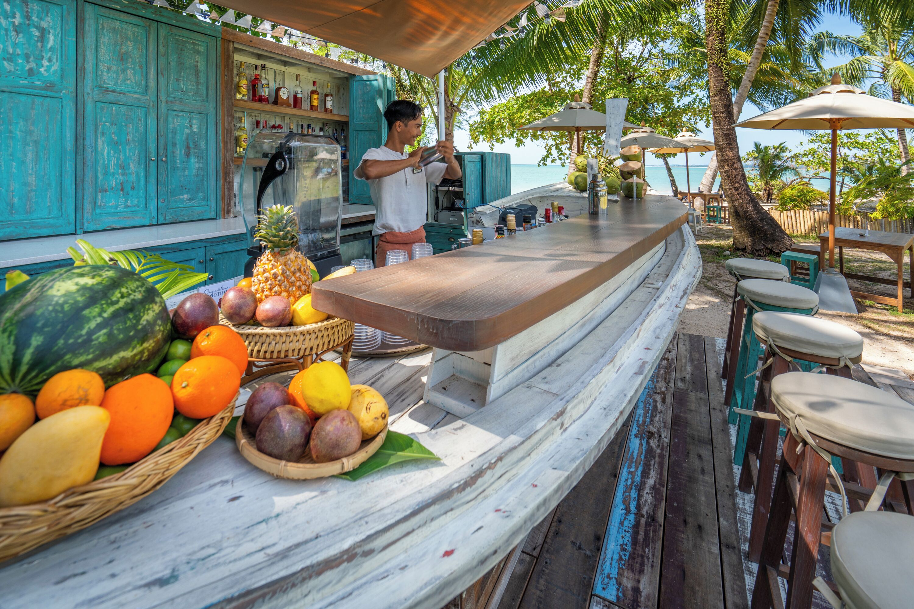Uma área de bar com balcão de madeira, exposição de frutas tropicais e vista para o mar.