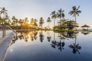 Piscina serena de borda infinita, com palmeiras e um pôr do sol refletido na água.