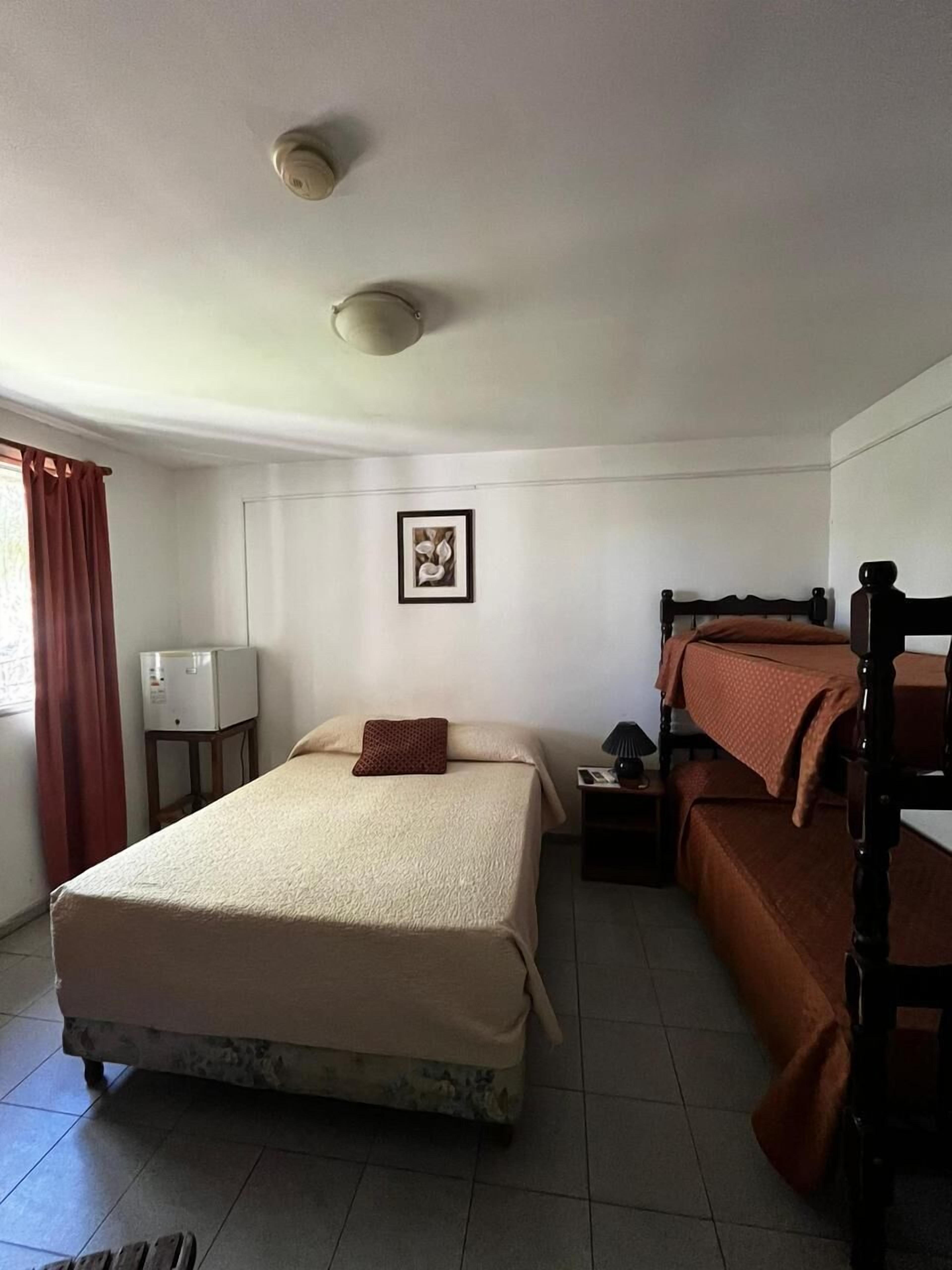 Uma cama de solteiro com cabeceira, mesa de cabeceira com abajur, quadro na parede e janela com cortinas.