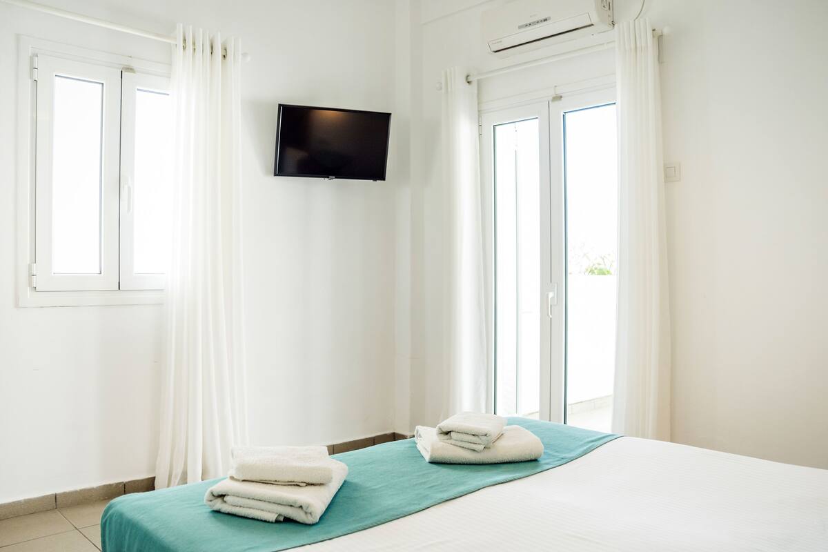 Quarto de hotel com uma cama, uma televisão fixada na parede e uma janela com cortinas.
