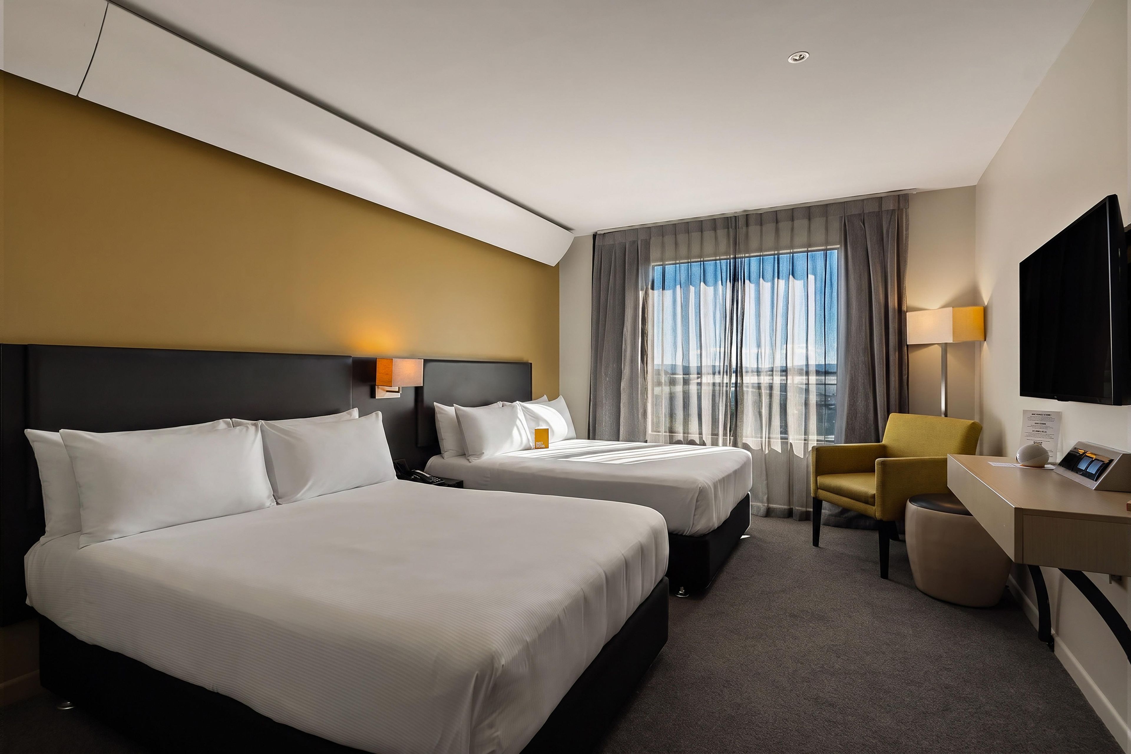 Quarto de hotel com duas camas, uma escrivaninha, uma cadeira, uma televisão e uma janela com cortinas.