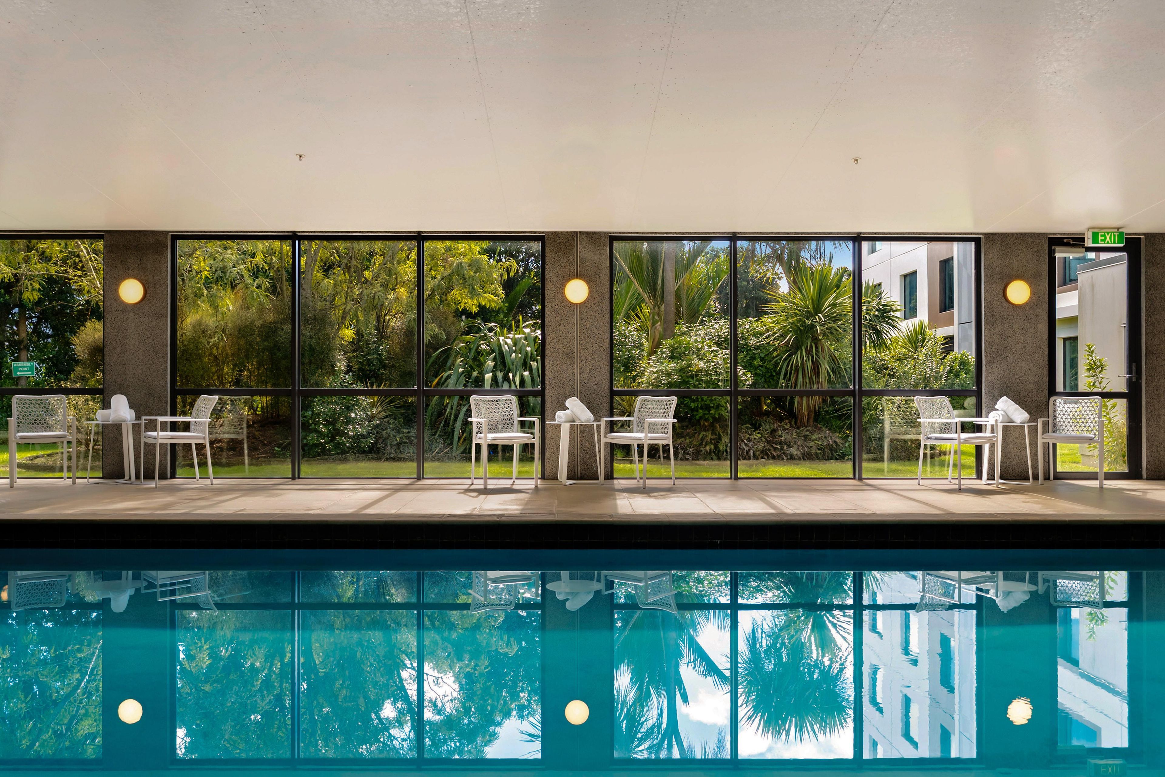 Área à beira da piscina com vista para um jardim através de grandes janelas, mobiliário de exterior e sinal de saída.