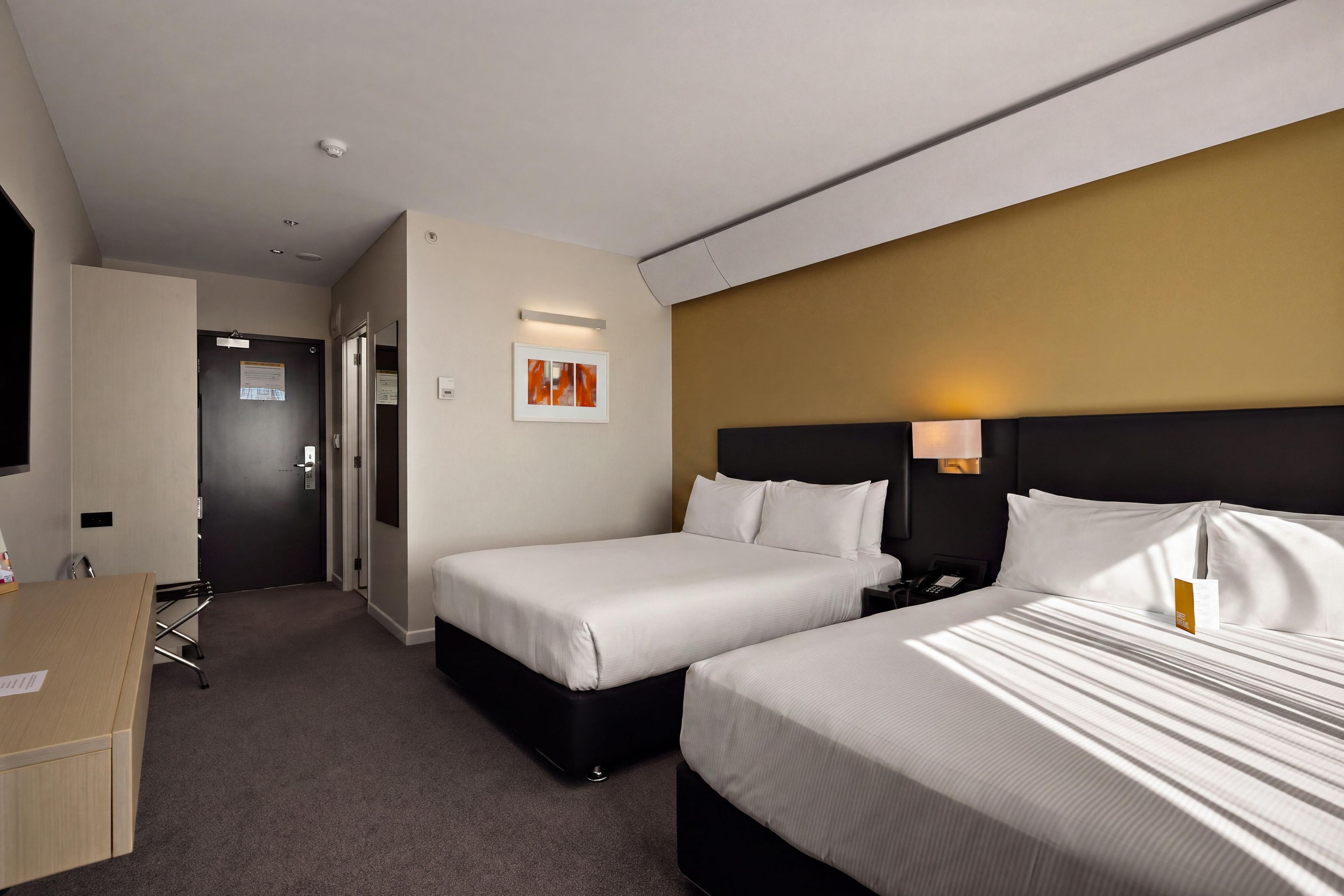 Quarto de hotel com duas camas, uma escrivaninha, uma cadeira, um guarda-roupa e uma porta.