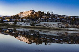 Um hotel com vários edifícios refletidos em águas calmas, rodeado por colinas e árvores cobertas de neve.