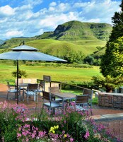 Um pátio com móveis de exterior, uma mesa e cadeiras, rodeado por flores e vista para uma paisagem verde com colinas.