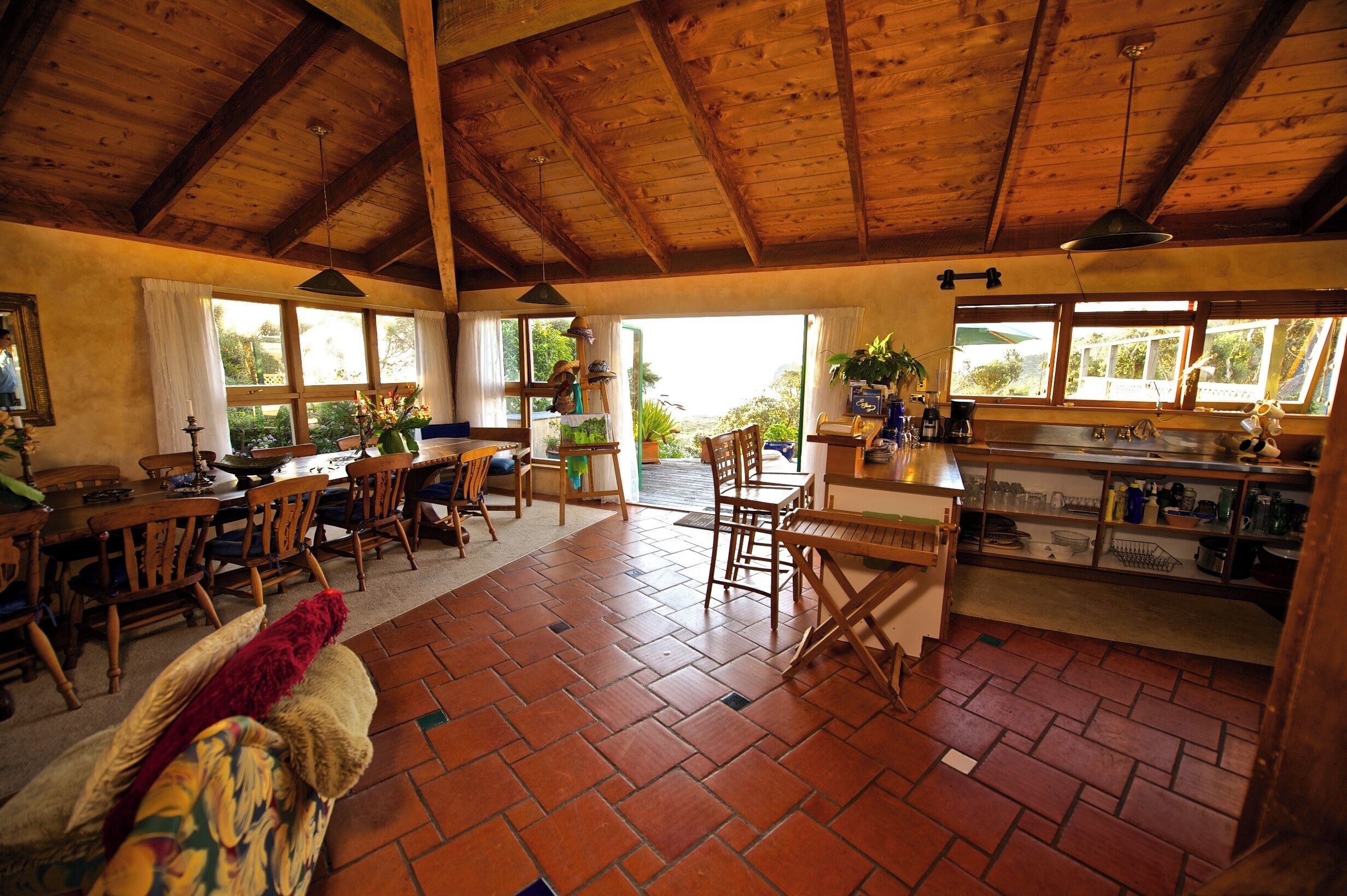 Ampla sala de estar com teto de madeira, mesa de jantar, cadeiras, sofá e cozinha com bancada e pia.