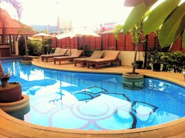 Área de piscina com espreguiçadeiras e guarda-sóis, cercada por plantas em vasos e um edifício vermelho ao fundo.
