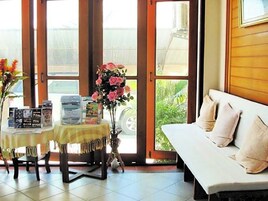 Quarto com sofá, mesa com revistas, vaso com flores e vista para o verde lá fora.
